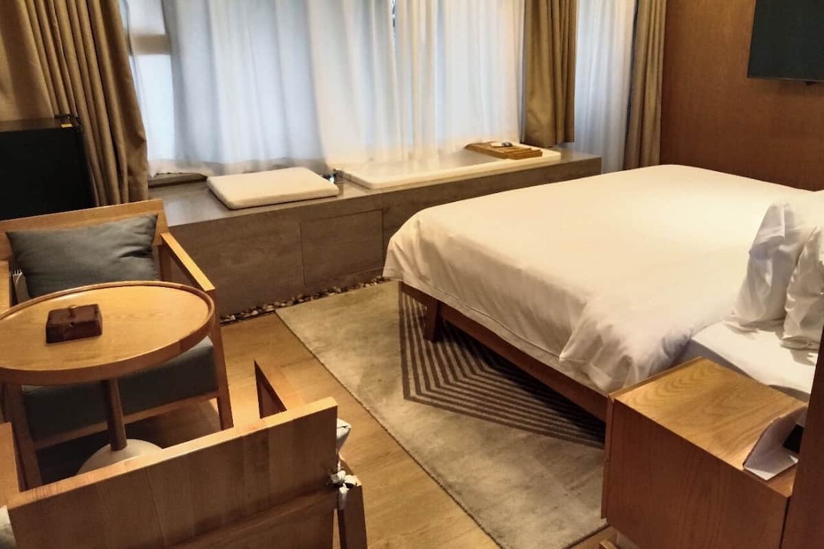 Quarto de hotel com uma cama, uma pequena mesa redonda com uma cadeira, um sofá e uma janela com cortinas.