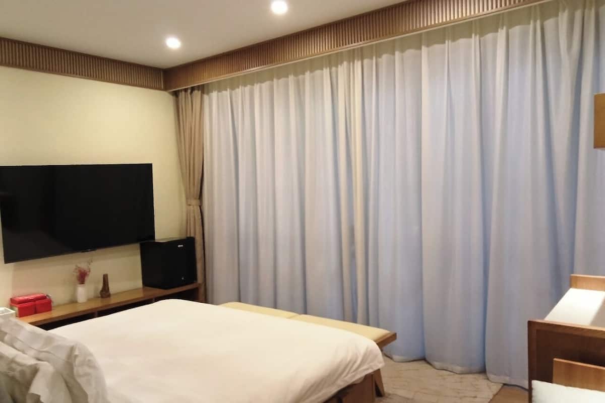 Quarto de hotel com cama, televisão presa à parede, mesa de cabeceira com alto-falante e uma janela grande com cortinas.