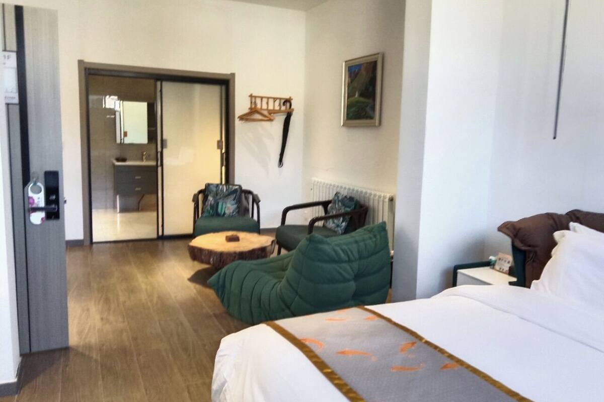 Quarto de hotel com uma cama, uma cadeira, uma mesinha e um sofá. Há uma porta que dá para um banheiro e um corredor.
