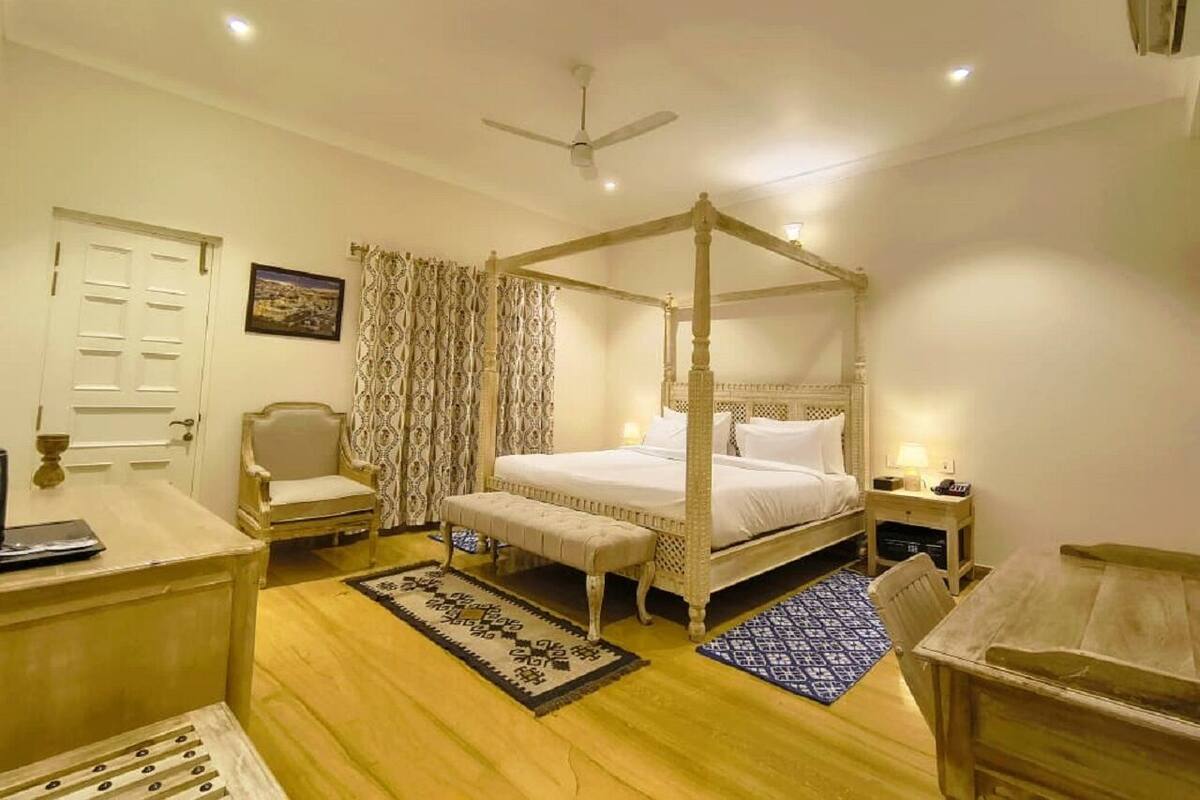 Uma cama com dossel e cortinas, mesa de cabeceira, cadeira, escrivaninha e pequena mesa com abajur.