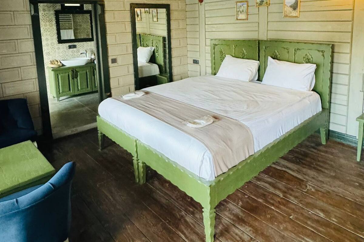 Quarto com piso de madeira, estrutura de cama verde, colcha branca, uma cadeira verde e um banheiro visível por uma porta aberta.