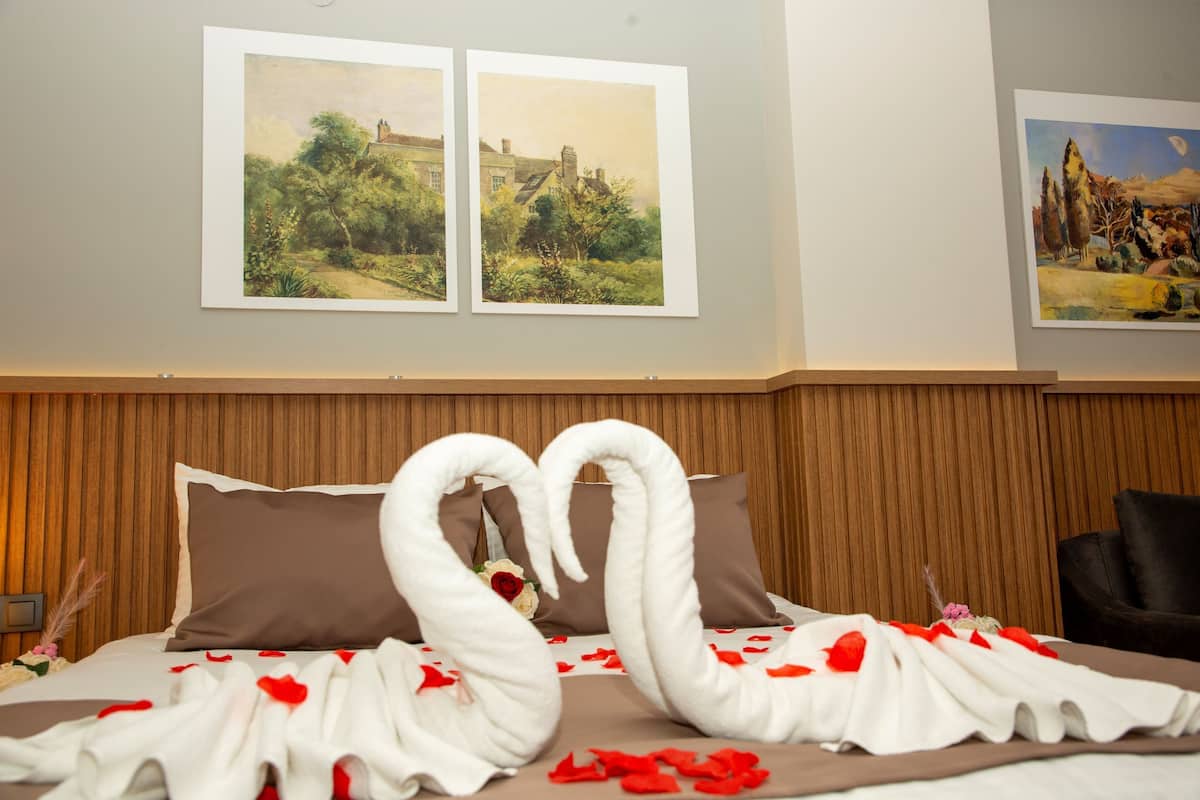 Quarto de hotel com cama decorada com almofadas em formato de cisne branco e pétalas de rosa vermelha, emoldurado por duas pinturas de paisagens.