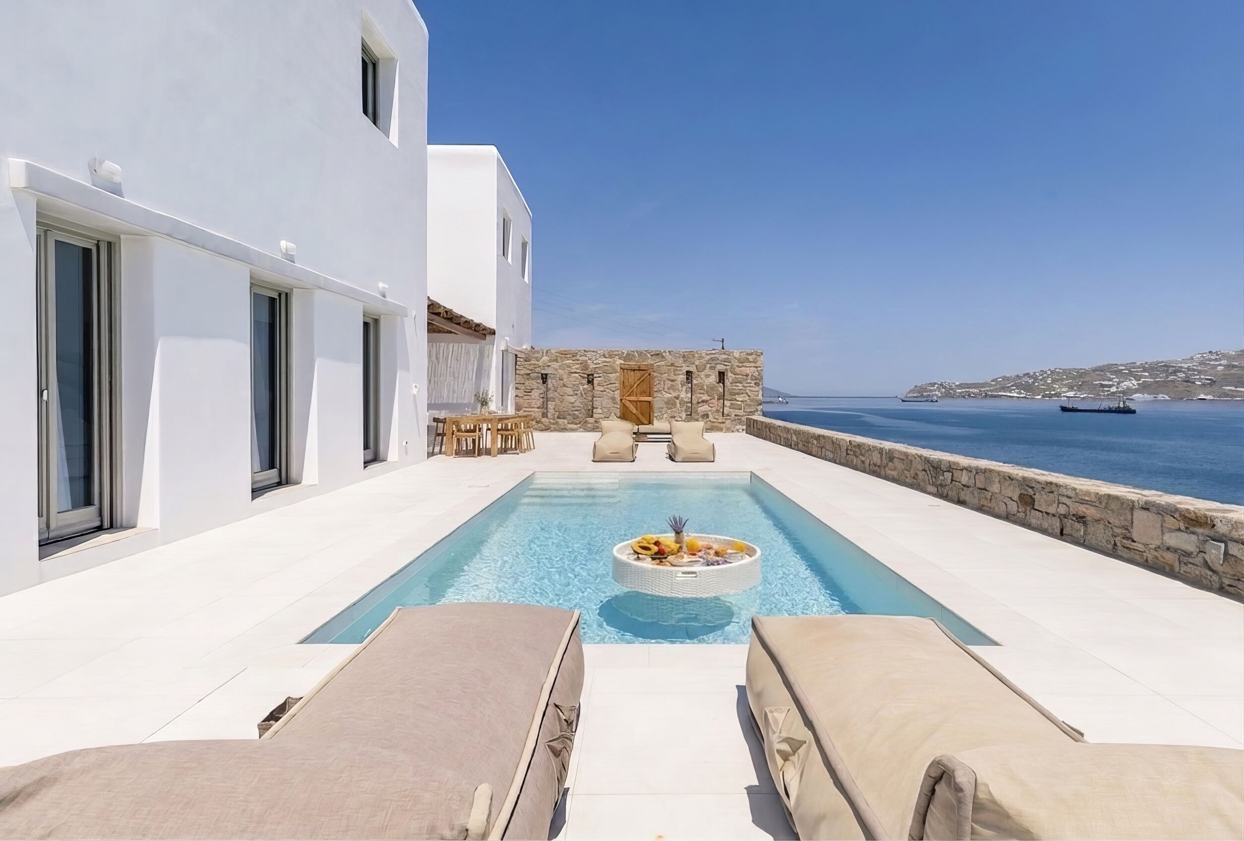 Uma área de piscina moderna com espreguiçadeiras, uma parede de pedra e vista para o mar.