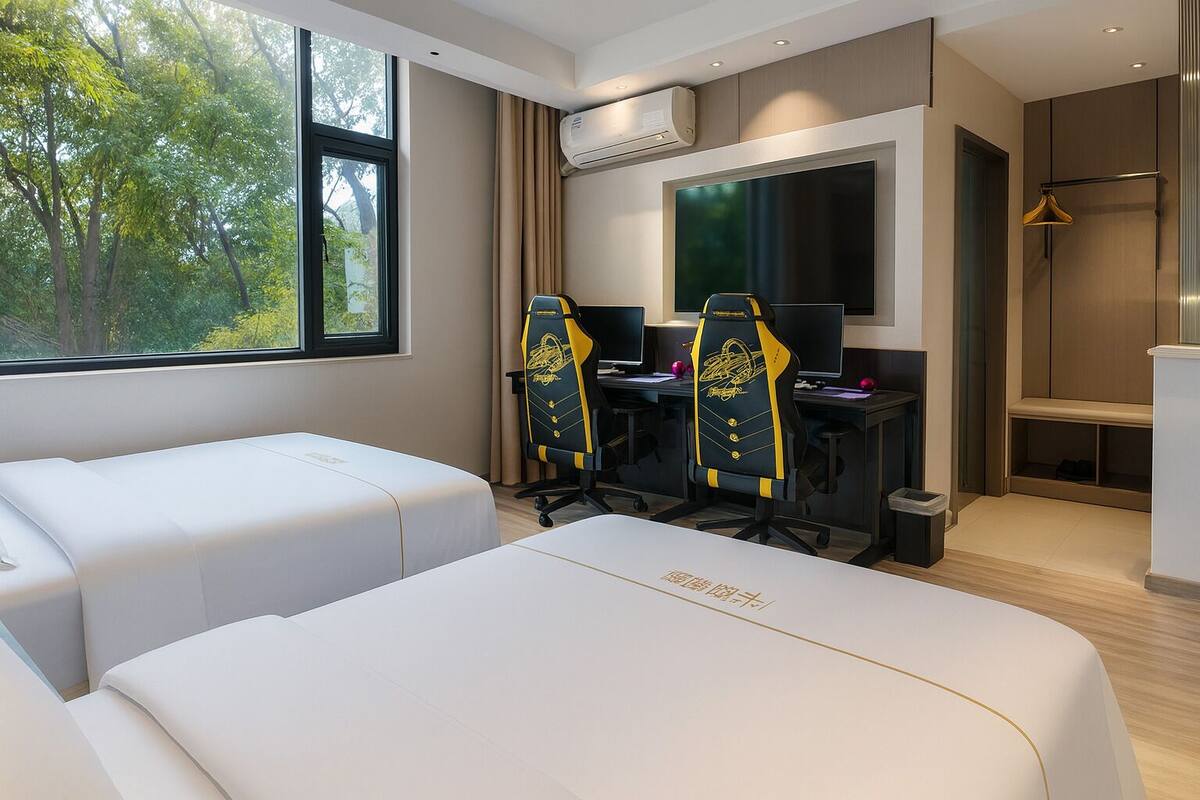 Quarto de hotel com duas camas, uma escrivaninha com dois computadores, uma televisão e um aparelho de ar condicionado.