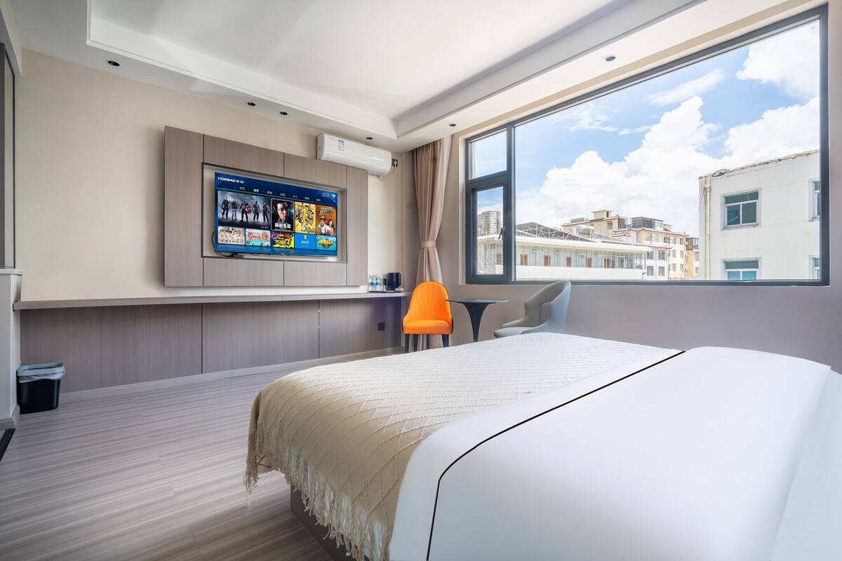 Quarto de hotel moderno com uma cama grande, uma televisão fixada na parede, uma janela com vista para a cidade e uma pequena mesa com uma cadeira.