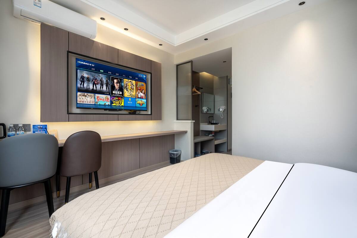 Quarto de hotel com cama, escrivaninha com duas cadeiras, TV fixada na parede exibindo cartazes de filmes, banheiro com espelho e pia, e um cesto de lixo.