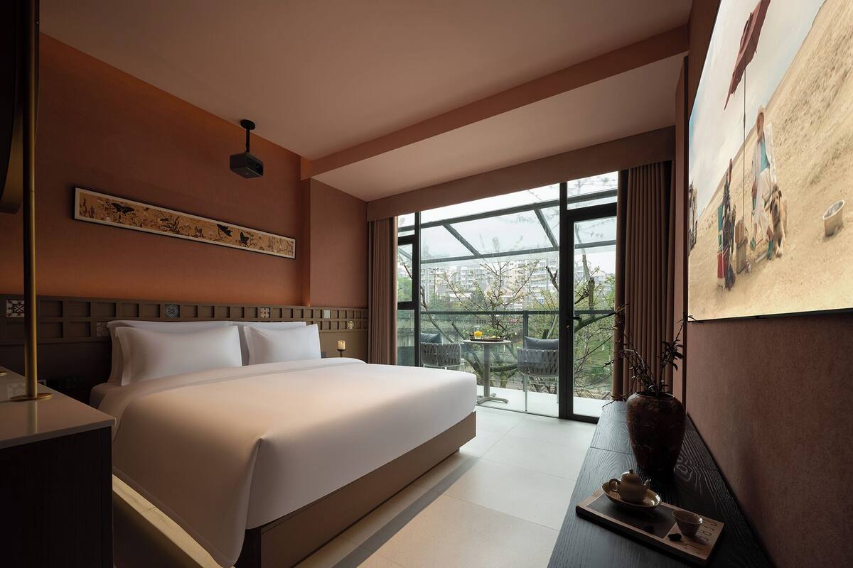 Quarto de hotel moderno com uma cama grande, uma mesa de trabalho e vista da cidade por grandes janelas.