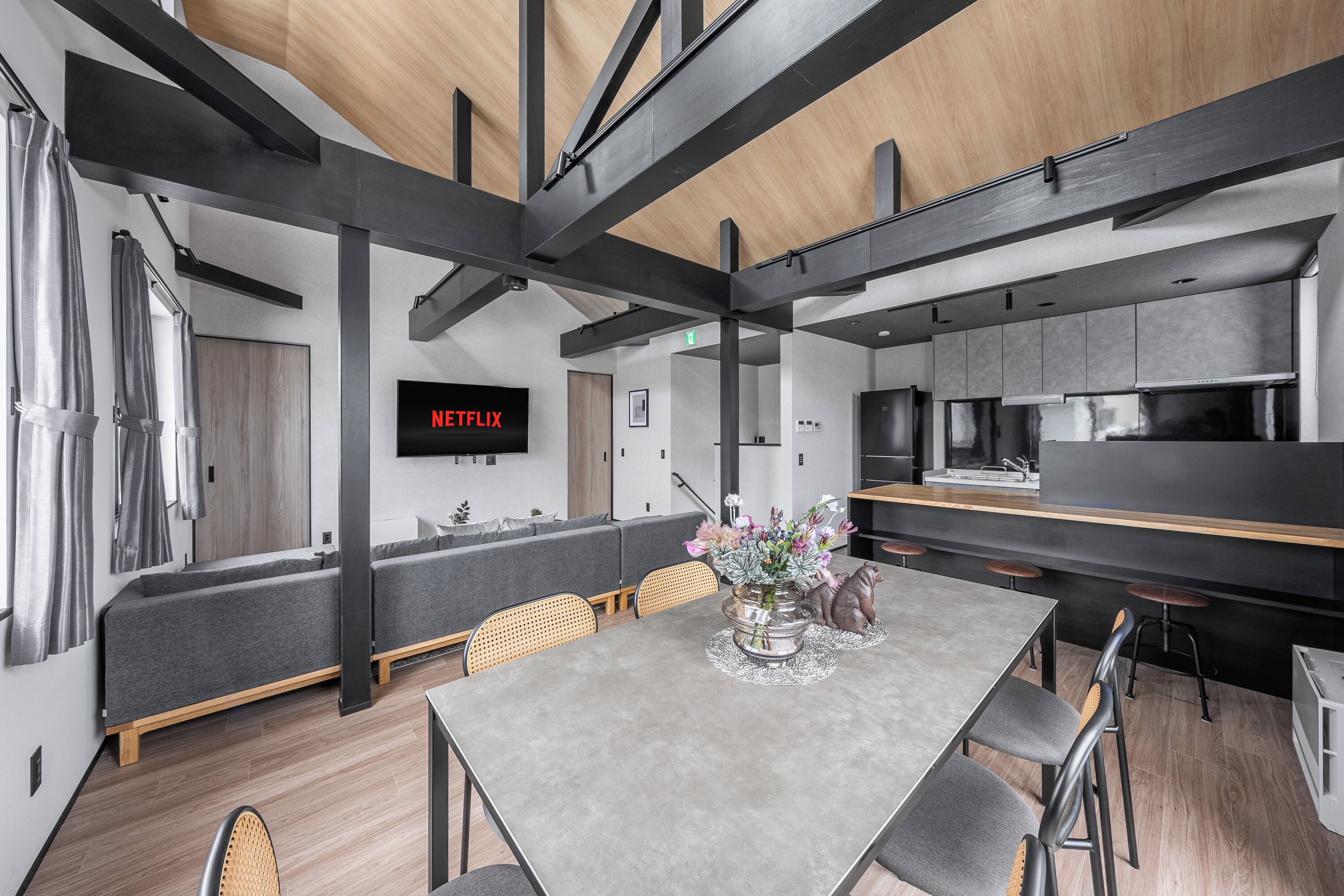 Uma cozinha moderna com forno e micro-ondas embutidos, uma área de jantar com mesa e cadeiras, e uma sala de estar com sofá e TV exibindo 'NETFLIX'.
