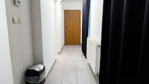 Um corredor estreito com uma porta de madeira no final, um radiador à direita e uma lixeira à esquerda.
