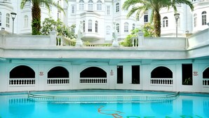 Uma piscina com 'AIGON PEARL' escrito na água, cercada por edifícios brancos com janelas em arco e palmeiras.