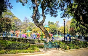 Um parque com equipamentos de playground, uma grande árvore e um banco.