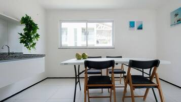 Uma cozinha moderna com bancada branca, duas cadeiras pretas, uma mesa branca e uma janela com vista para um edifício.