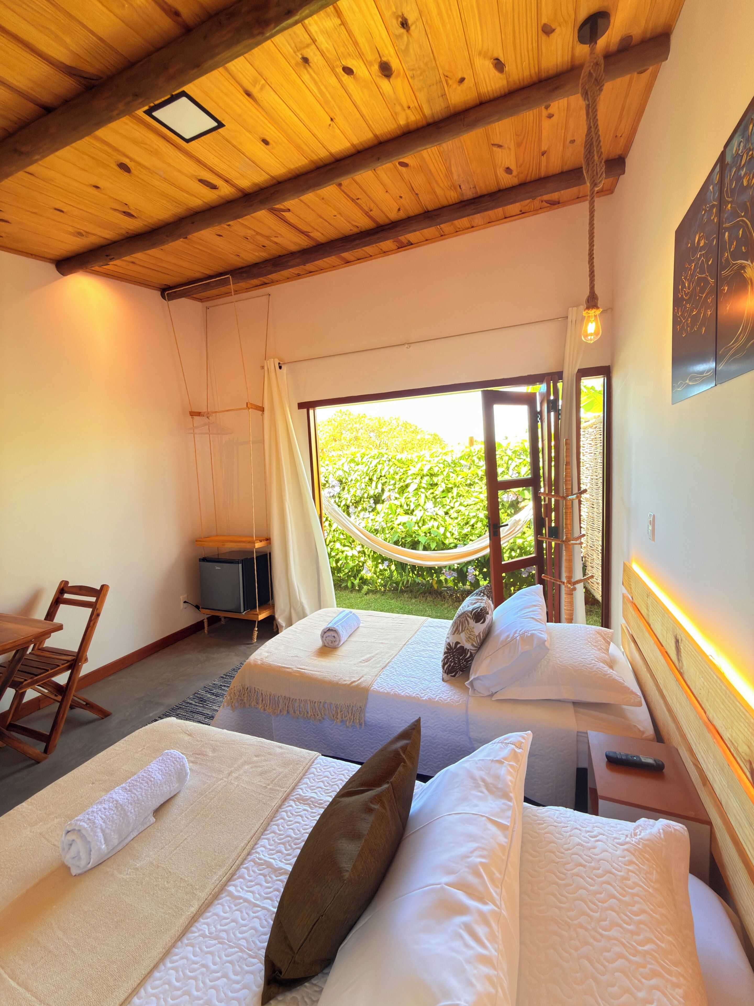 Quarto com duas camas, teto de madeira, janela com vista para o verde e uma pequena mesa.