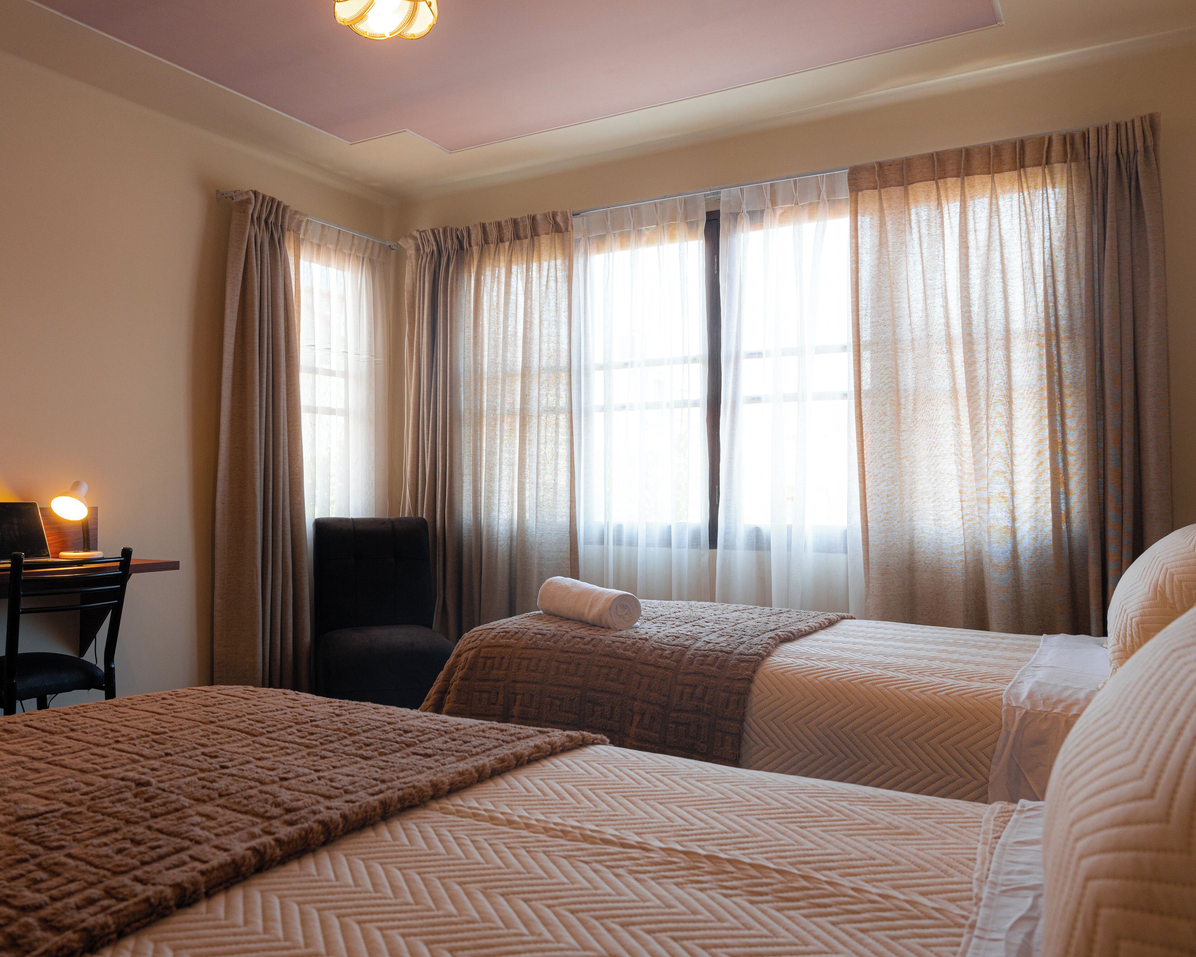 Quarto de hotel com duas camas, uma mesa com luminária e uma cadeira.