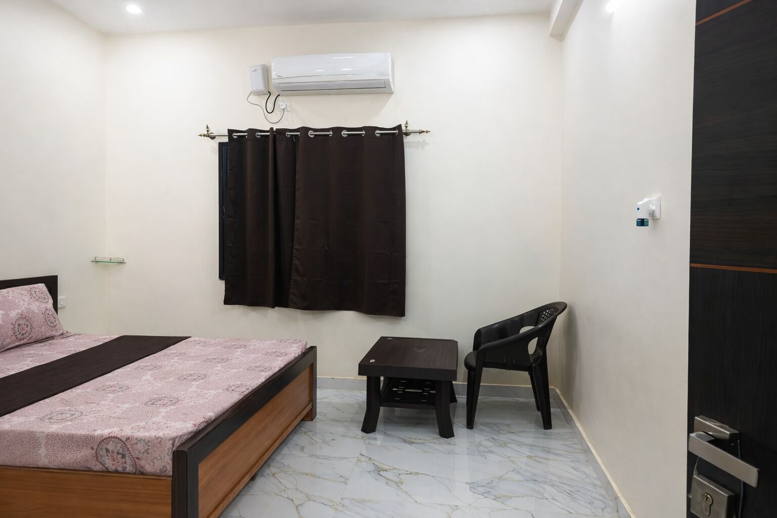 Quarto com cama, mesa pequena, cadeira, ar condicionado e guarda-roupa escuro.