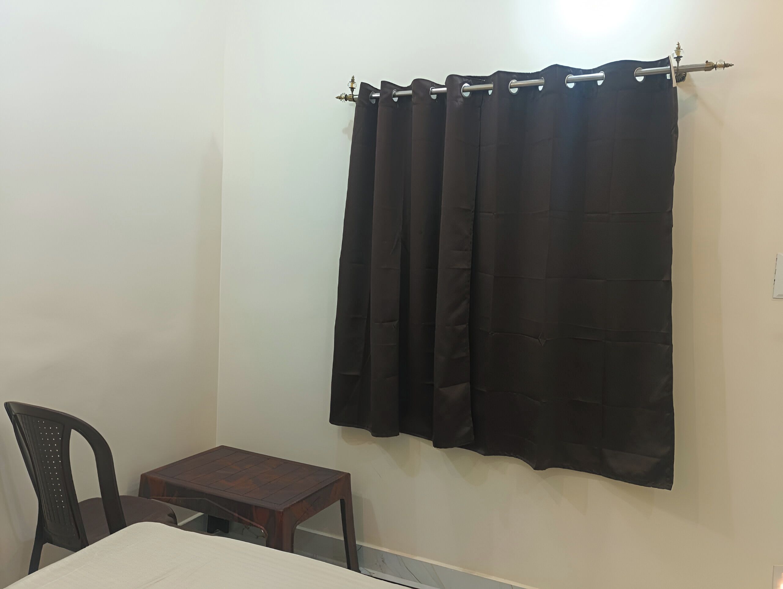 Um quarto pequeno com uma cama, uma mesa de madeira, uma cadeira preta e uma cortina escura na janela.