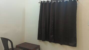Um quarto pequeno com uma cama, uma mesa de madeira, uma cadeira preta e uma cortina escura na janela.