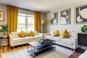 Uma sala de estar moderna com dois sofás brancos, uma mesa de centro preta e almofadas amarelas. Há quadros abstratos na parede e um abajur.