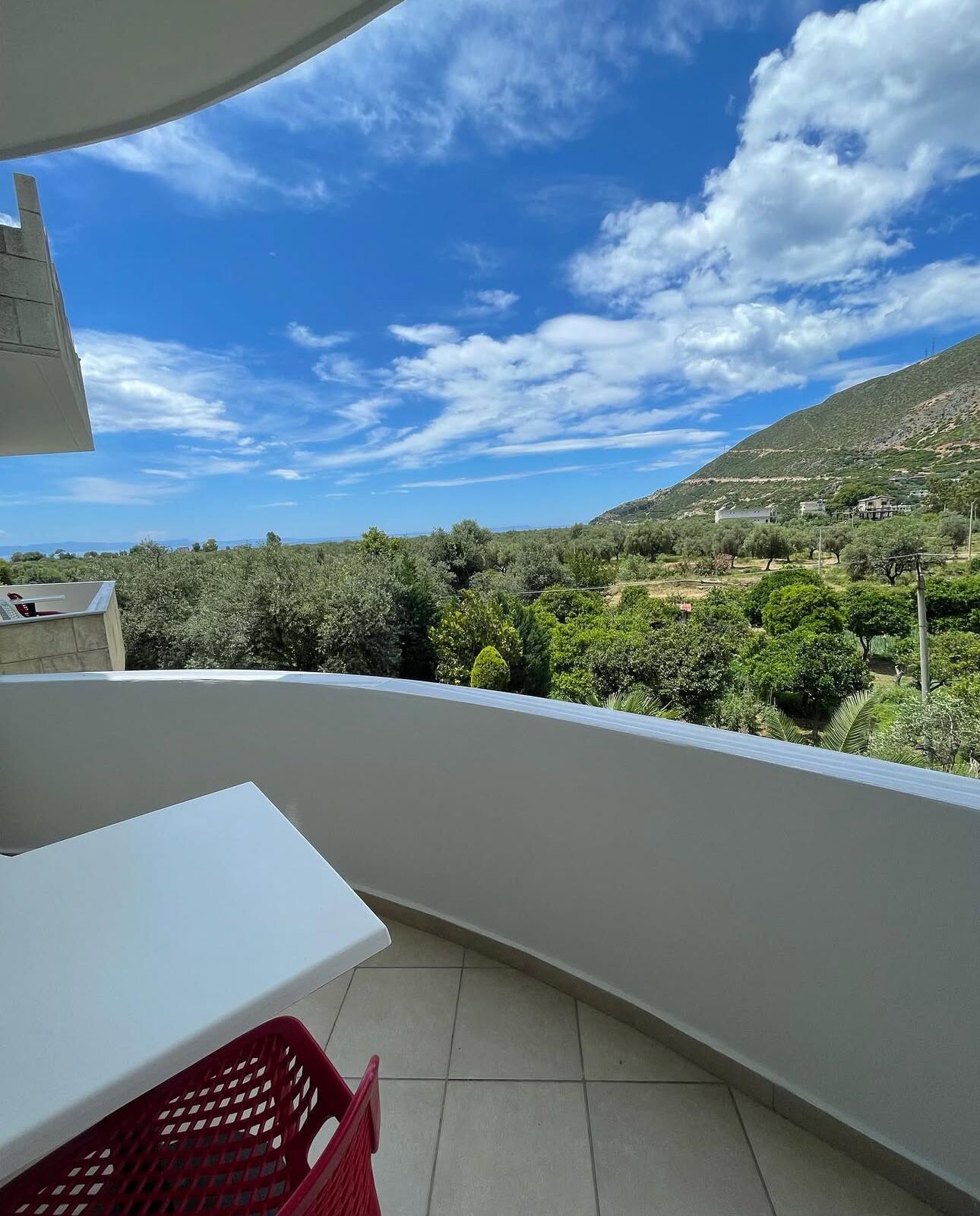 Uma varanda com mesa branca e cadeira vermelha, com vista para uma paisagem verde com árvores e uma colina.
