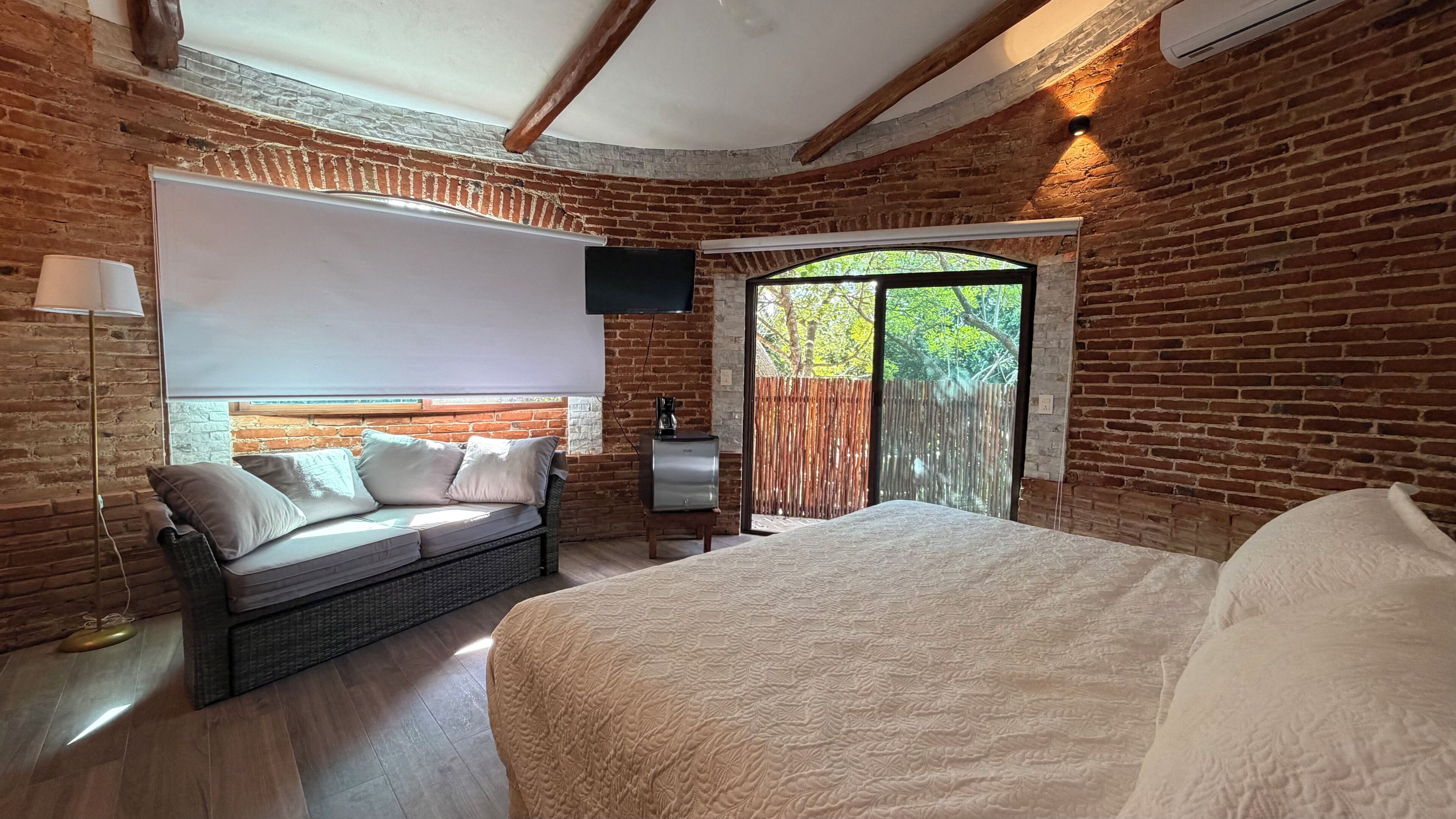 Quarto com cama, sofá, televisão e janela com vista para vegetação.
