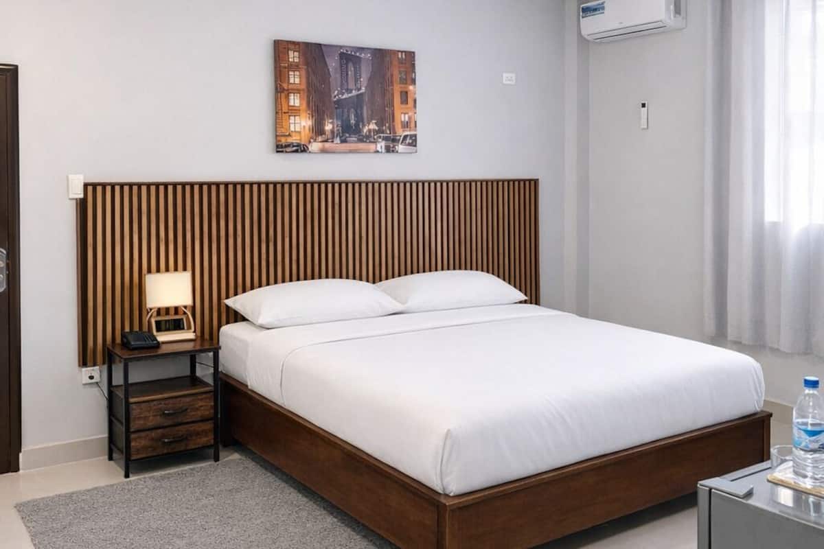 Quarto de hotel moderno com cabeceira de madeira, cama com roupa de cama branca, mesa de cabeceira com abajur, um quadro na parede e um ar condicionado.