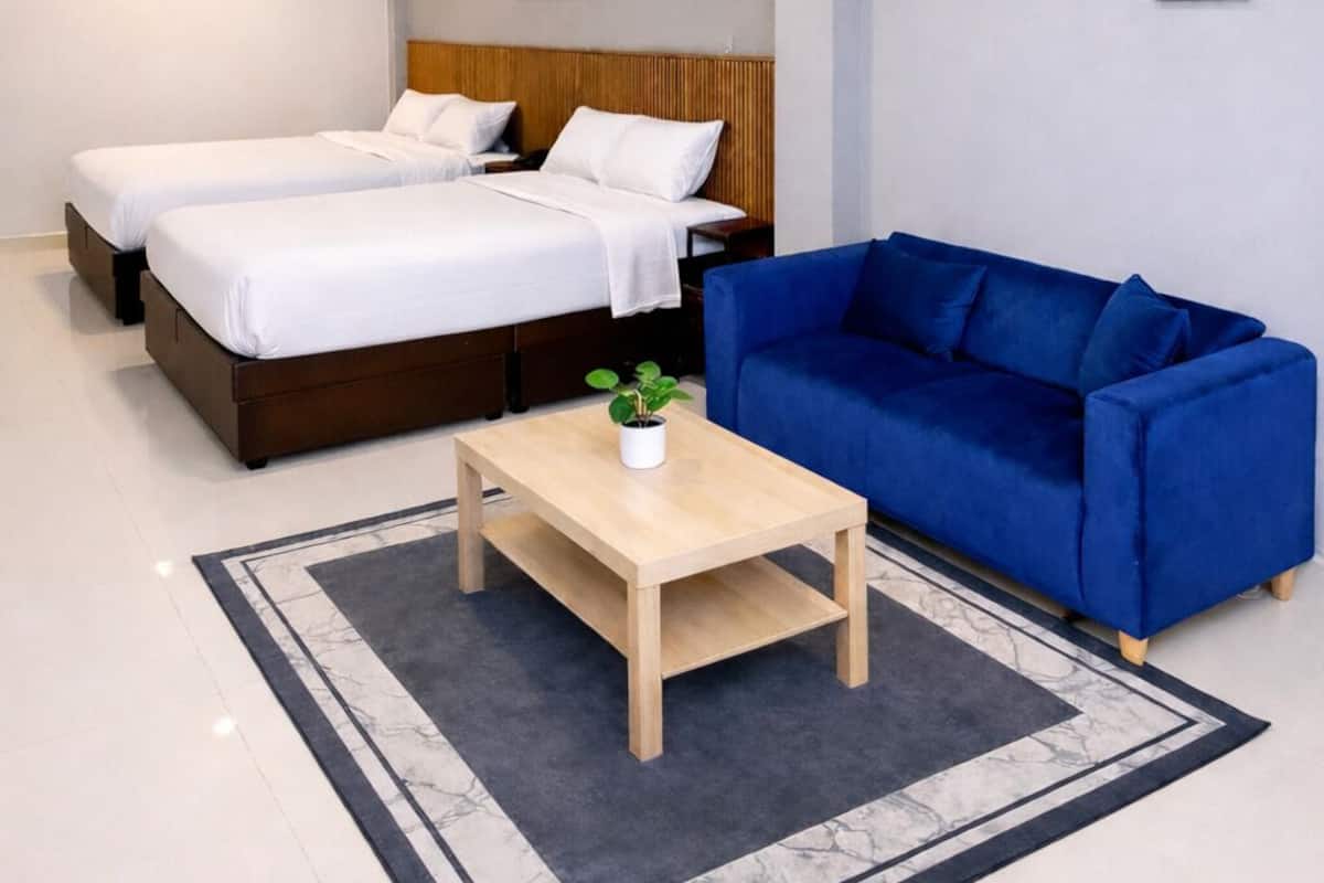 Quarto de hotel com duas camas, um sofá azul, uma mesa de centro de madeira e uma pequena planta.