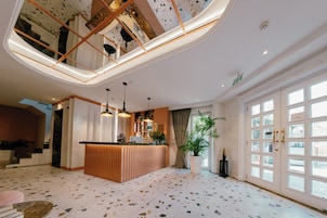 Um saguão de hotel com balcão de recepção, luminárias pendentes, uma janela ampla e piso de terrazzo.