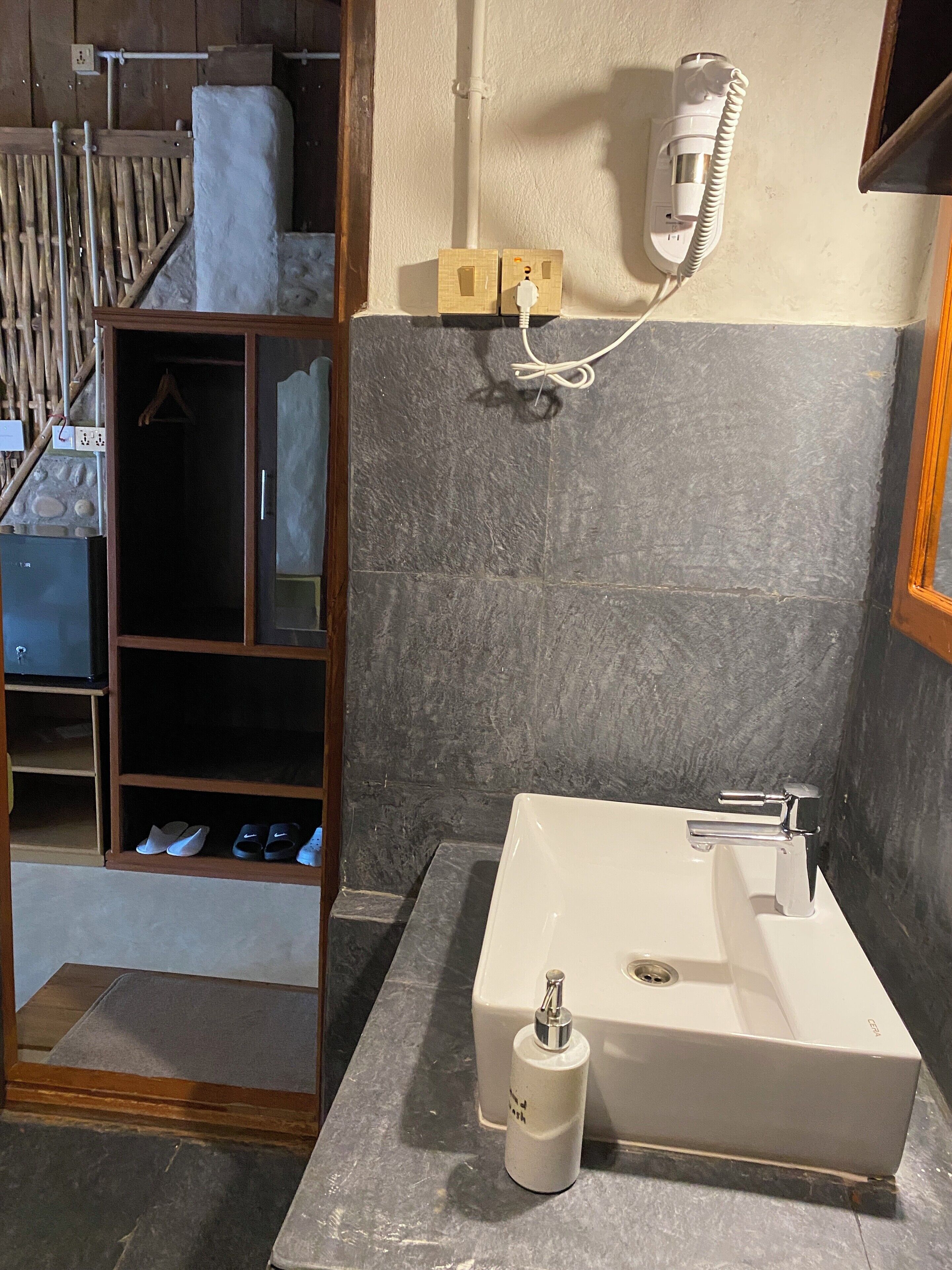 Casa de banho com lavatório branco, uma prateleira de madeira com sapatos, uma toalha e um telefone fixado na parede.