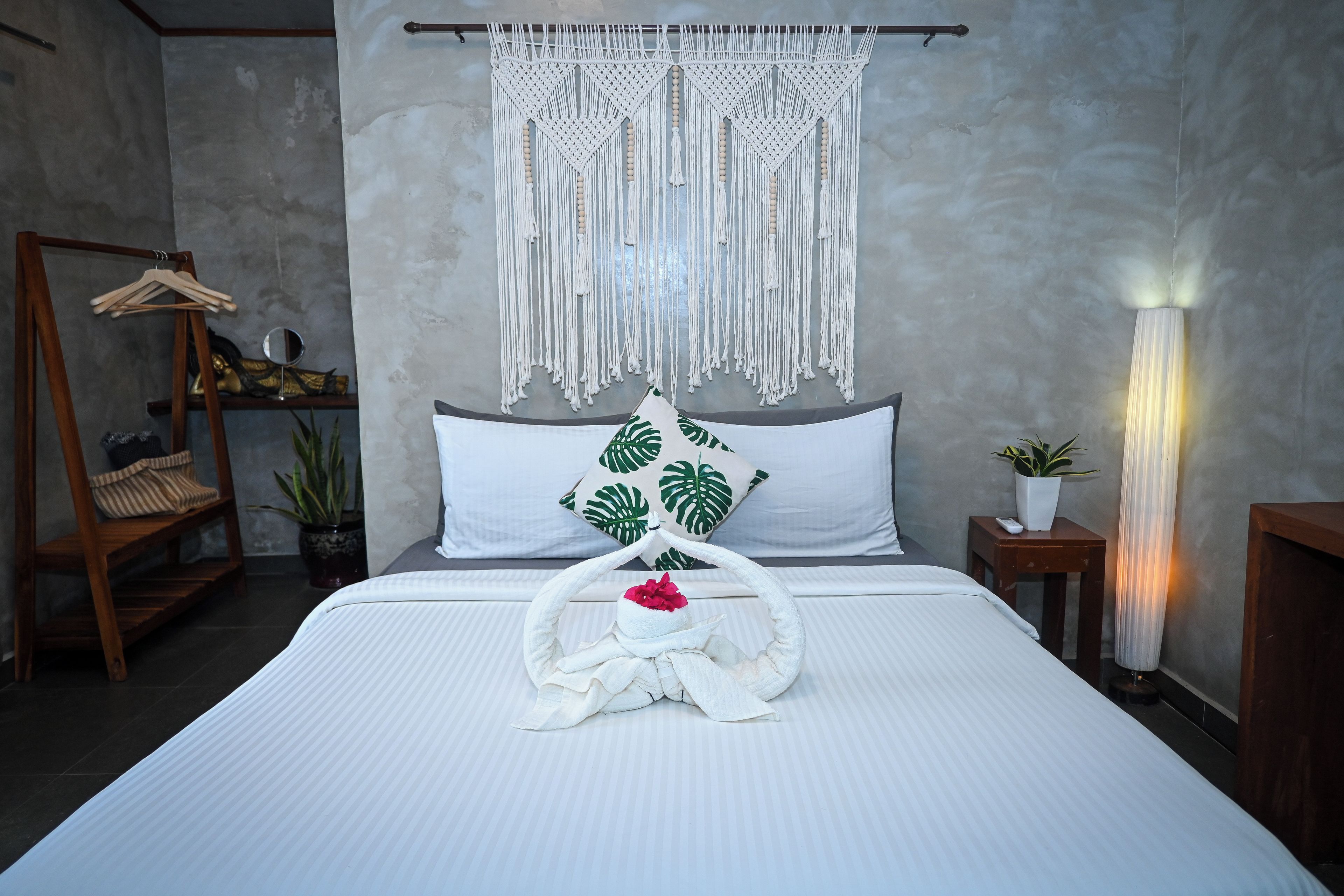 Cama bem arrumada com um pendente de macramé, um cabideiro de madeira e um abajur.