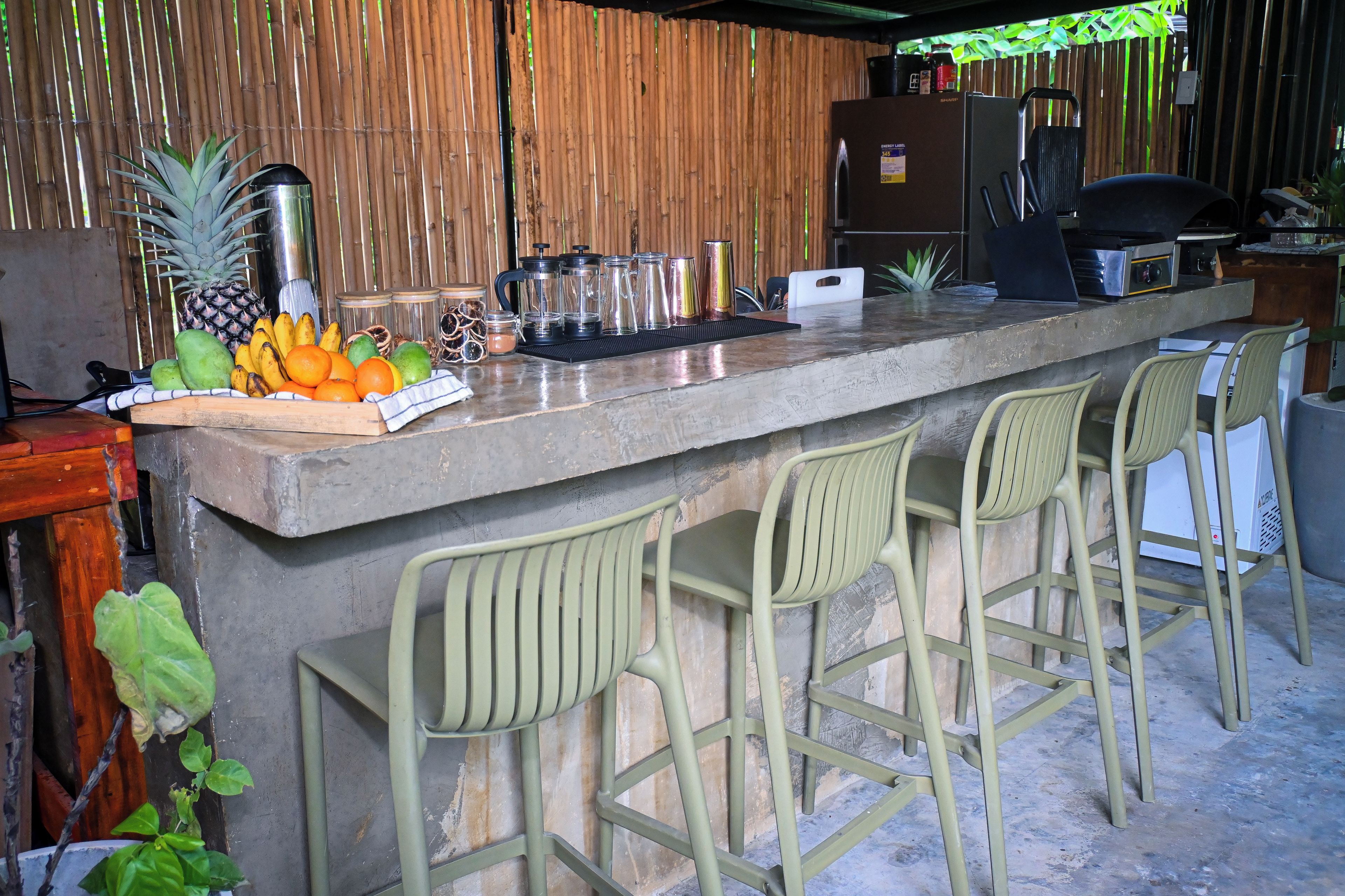 Uma área de bar com bancada de concreto, cafeteira, liquidificador e geladeira. Três cadeiras verdes e uma variedade de frutas e bebidas estão sobre a bancada.