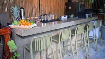 Uma área de bar com bancada de concreto, cafeteira, liquidificador e geladeira. Três cadeiras verdes e uma variedade de frutas e bebidas estão sobre a bancada.