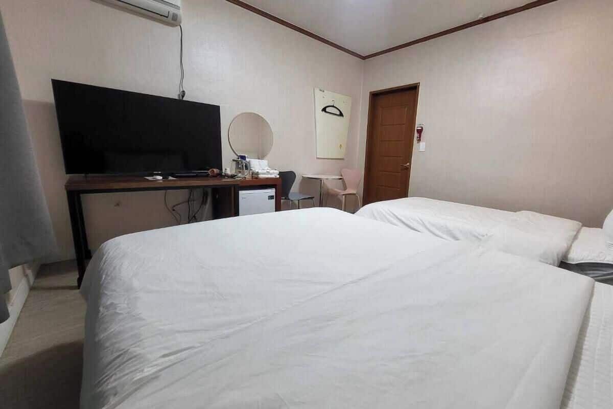 Quarto de hotel com duas camas, uma mesa, uma televisão de tela plana, uma mesinha, uma cadeira e uma porta.