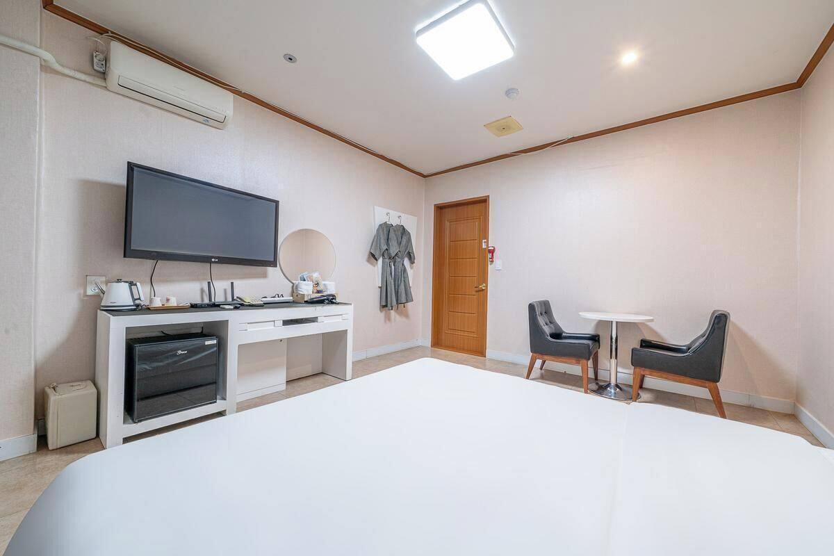 Quarto de hotel com uma cama grande, uma escrivaninha com televisão de tela plana, uma mesinha, duas cadeiras, um cabideiro e uma porta.