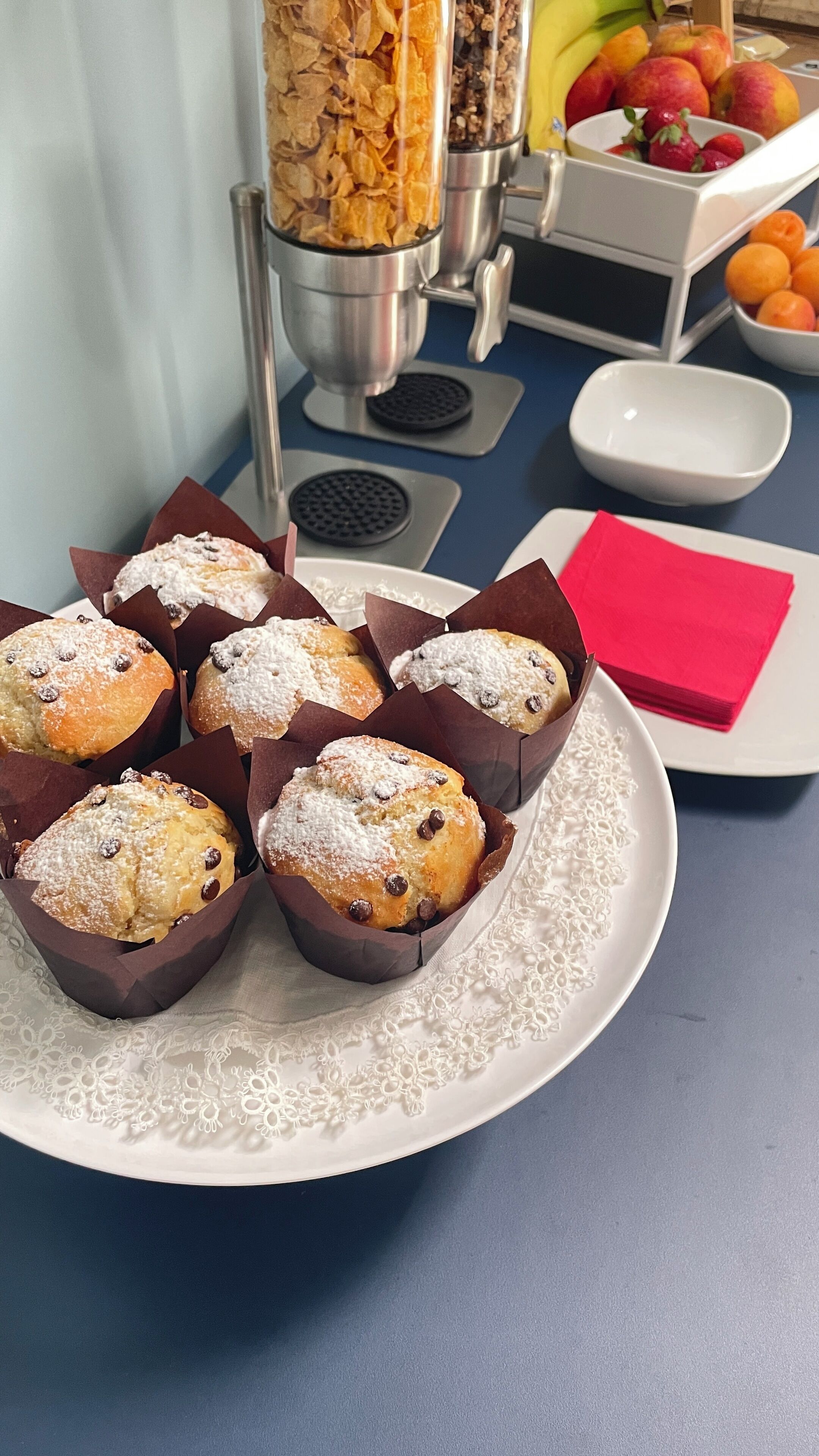 Um prato de muffins com açúcar de confeiteiro e gotas de chocolate, uma cafeteira e uma fruteira sobre uma mesa.
