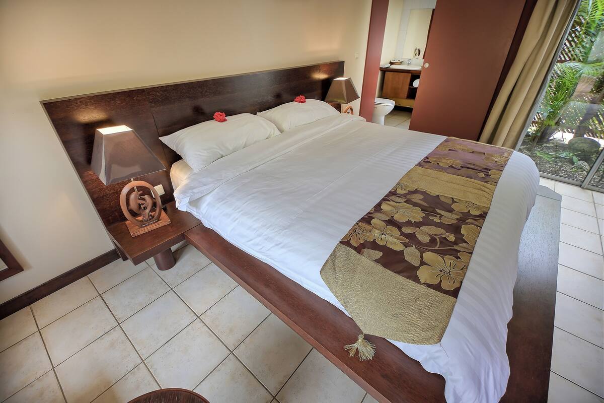 Quarto de hotel com uma cama grande, uma mesa de cabeceira com abajur, uma pequena mesa com um objeto decorativo e vista para o exterior pela janela.
