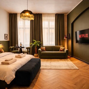 Quarto de hotel com uma cama, um sofá, uma televisão e uma janela grande com cortinas.