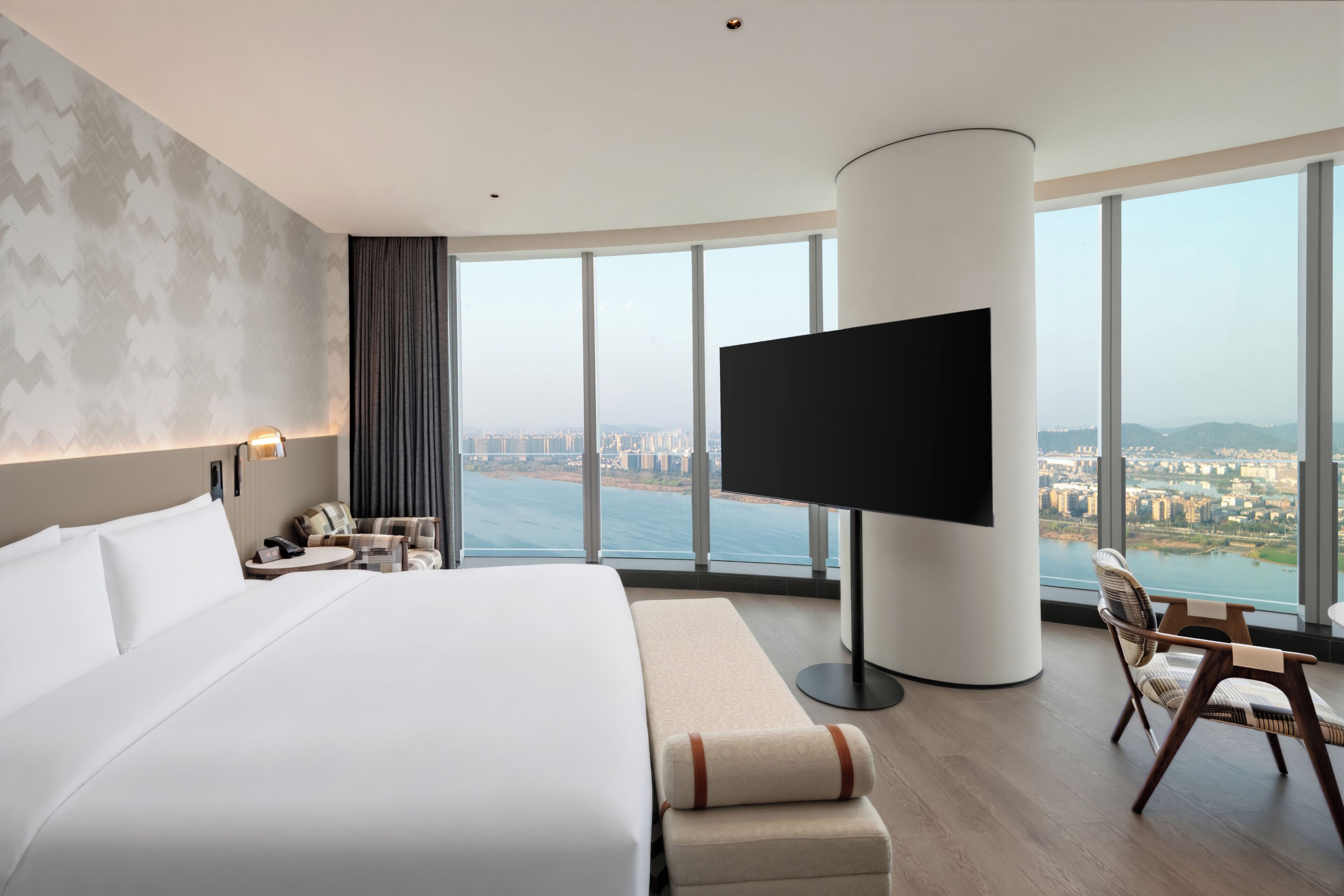 Quarto de hotel moderno com uma cama grande, televisão de tela plana, área de estar e vista para o horizonte urbano através de amplas janelas.