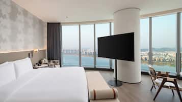 Quarto de hotel moderno com uma cama grande, televisão de tela plana, área de estar e vista para o horizonte urbano através de amplas janelas.