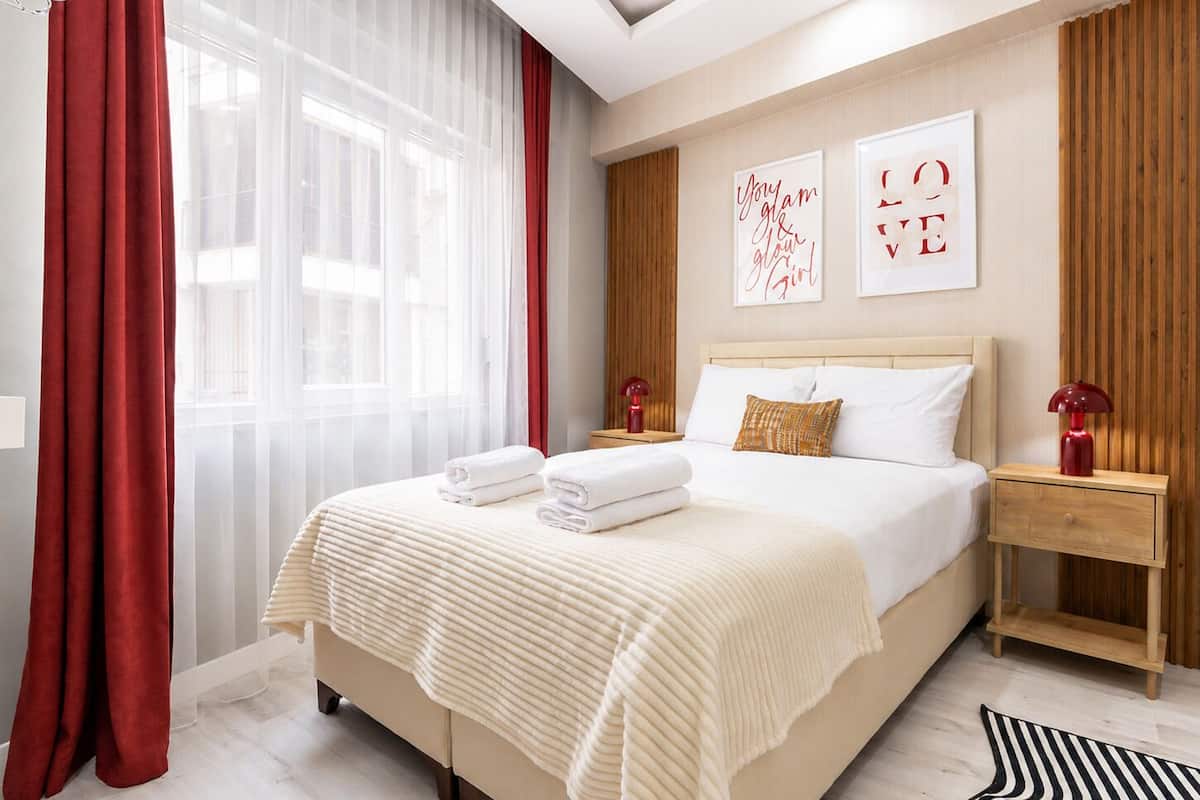 Quarto de hotel com cama, duas mesas de cabeceira, uma mesinha de apoio, janela com cortinas leves e obras de arte na parede.