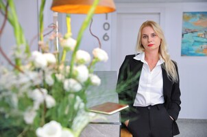 Uma mulher com um blazer preto e camisa branca sentada à mesa com um livro, uma luminária e um vaso de flores.