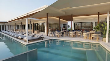 Uma área à beira da piscina com espreguiçadeiras e mesas, piscina cristalina e um espaço de estar coberto com grandes janelas.