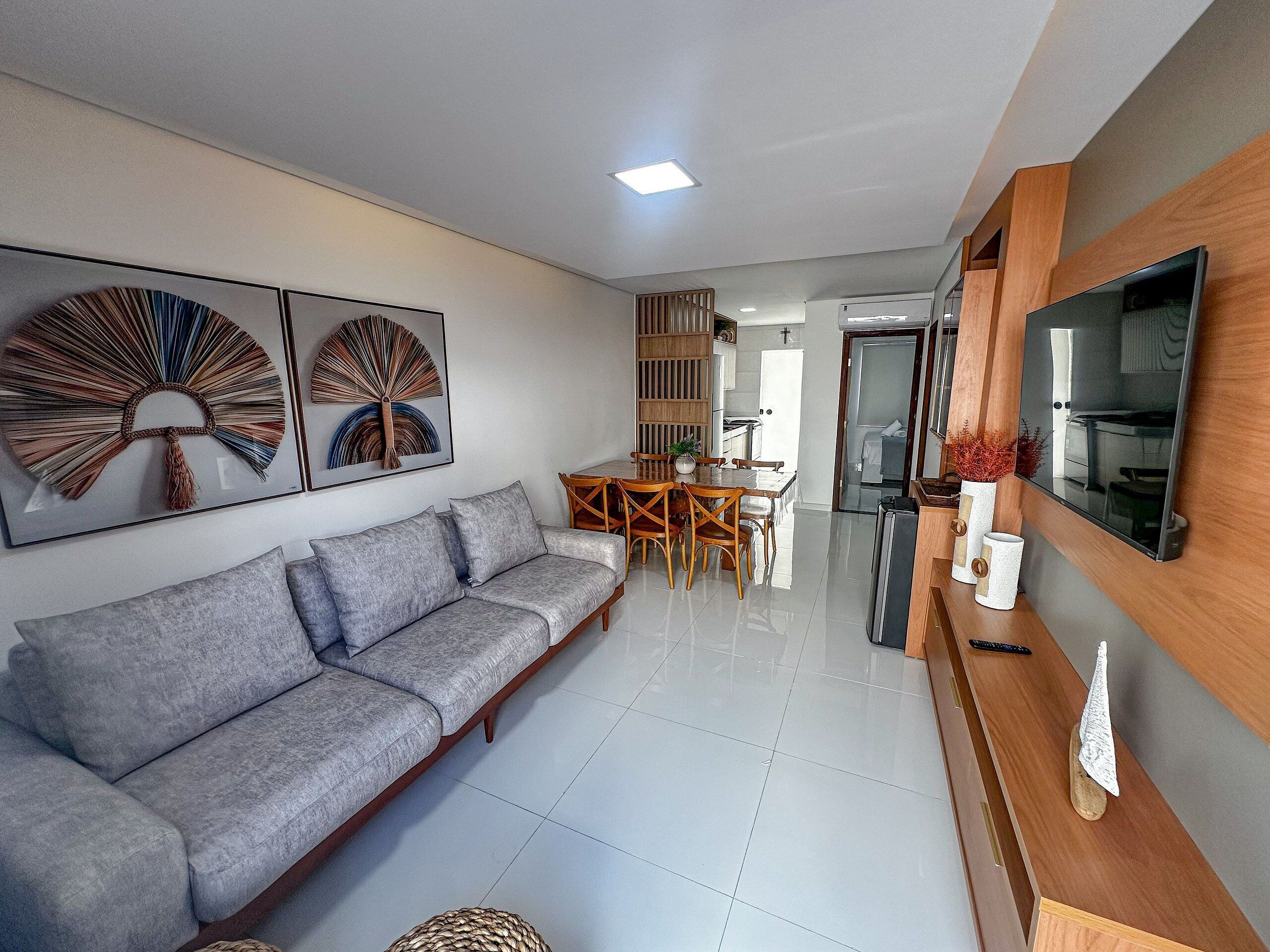 Uma sala de estar moderna com um sofá cinzento, mesa de jantar de madeira e televisão fixada na parede.