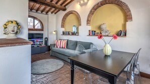 Uma sala de estar aconchegante com teto de madeira, sofá cinza, mesa de jantar com cadeiras e uma TV ao fundo.