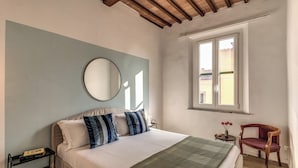 Quarto com cama, mesa de cabeceira com abajur, cadeira e janela.