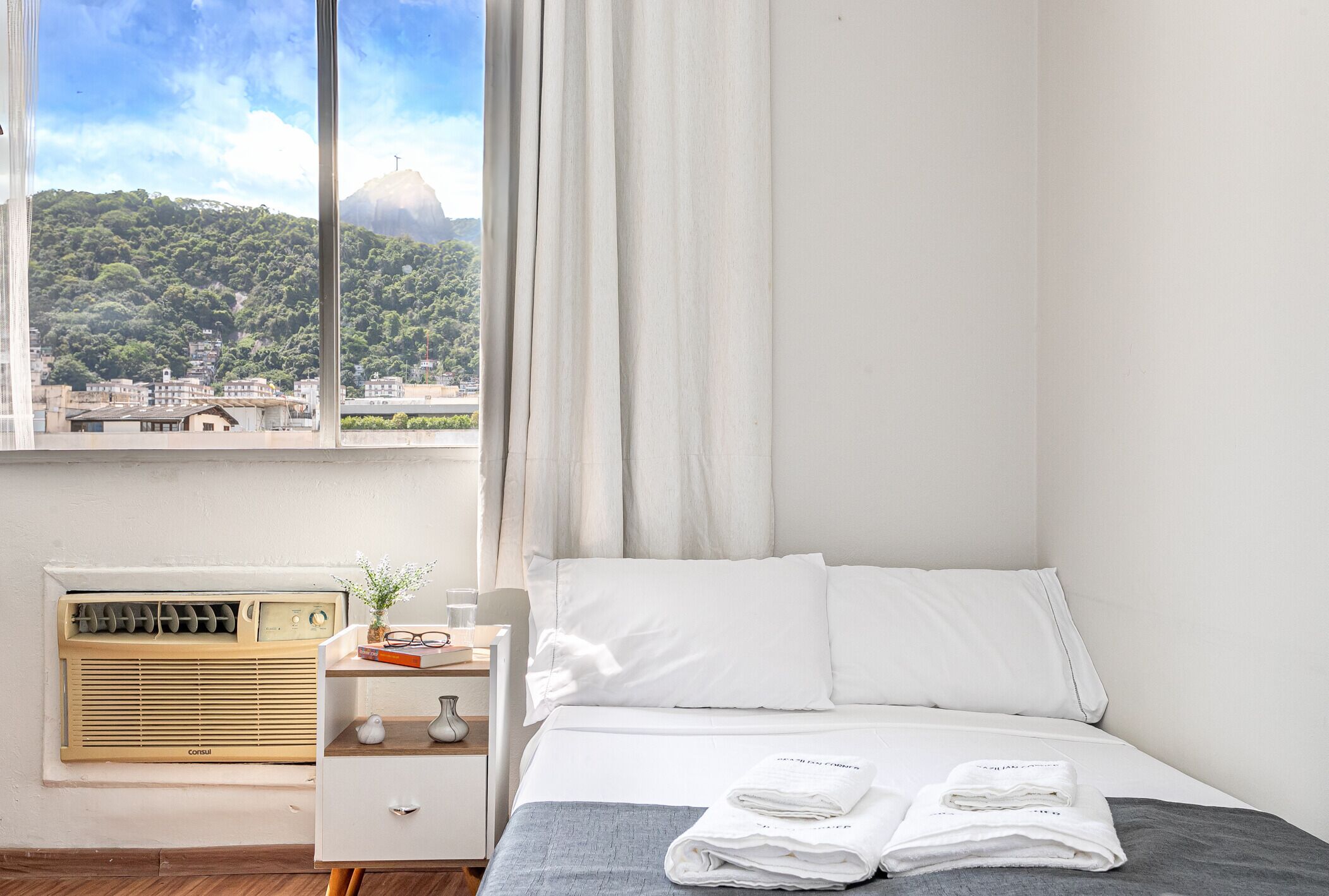 Quarto de hotel com cama, janela com vista para uma montanha, radiador e mesa de cabeceira com um vaso e copos.
