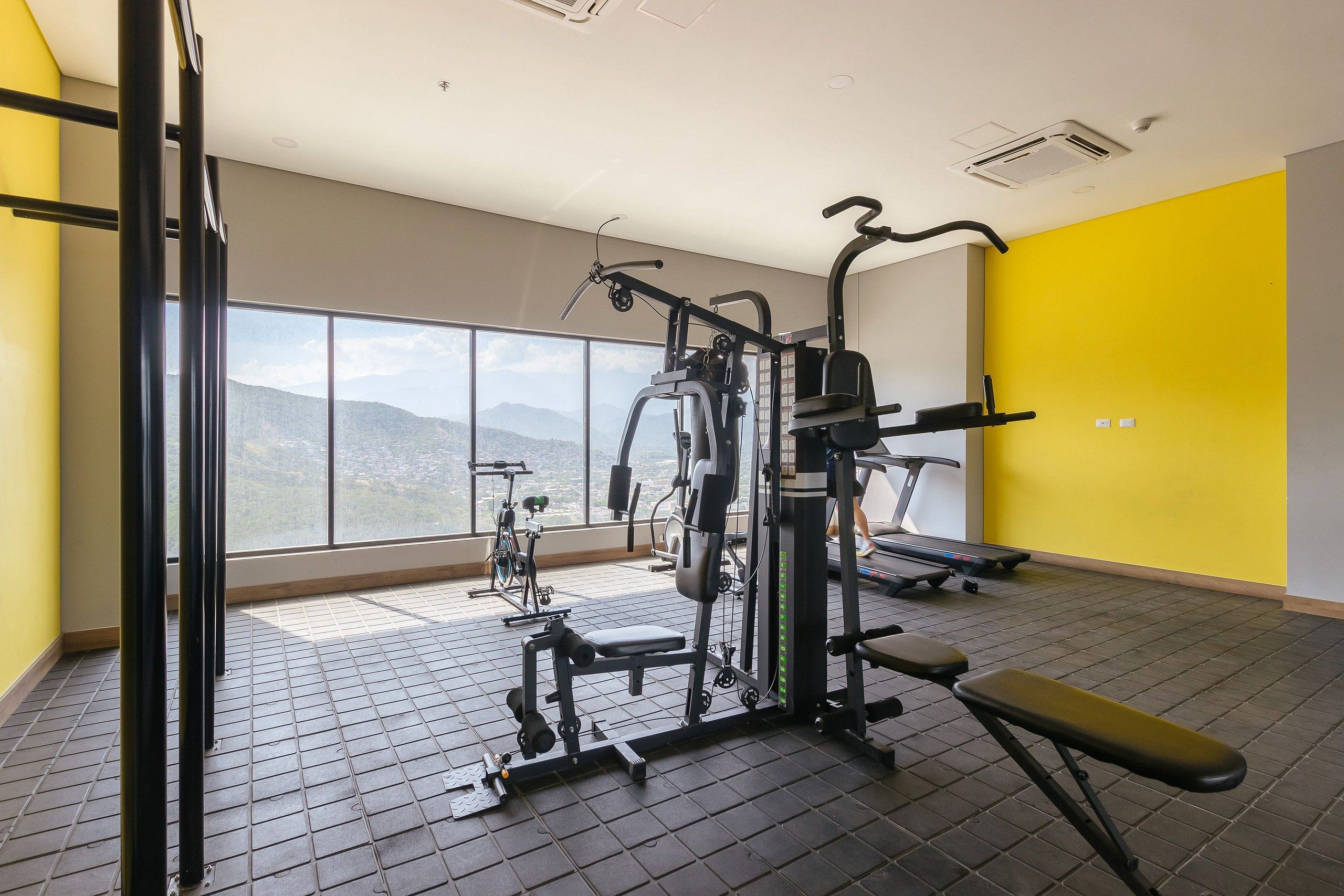 Uma academia com equipamentos de exercícios, grandes janelas e uma parede amarela.