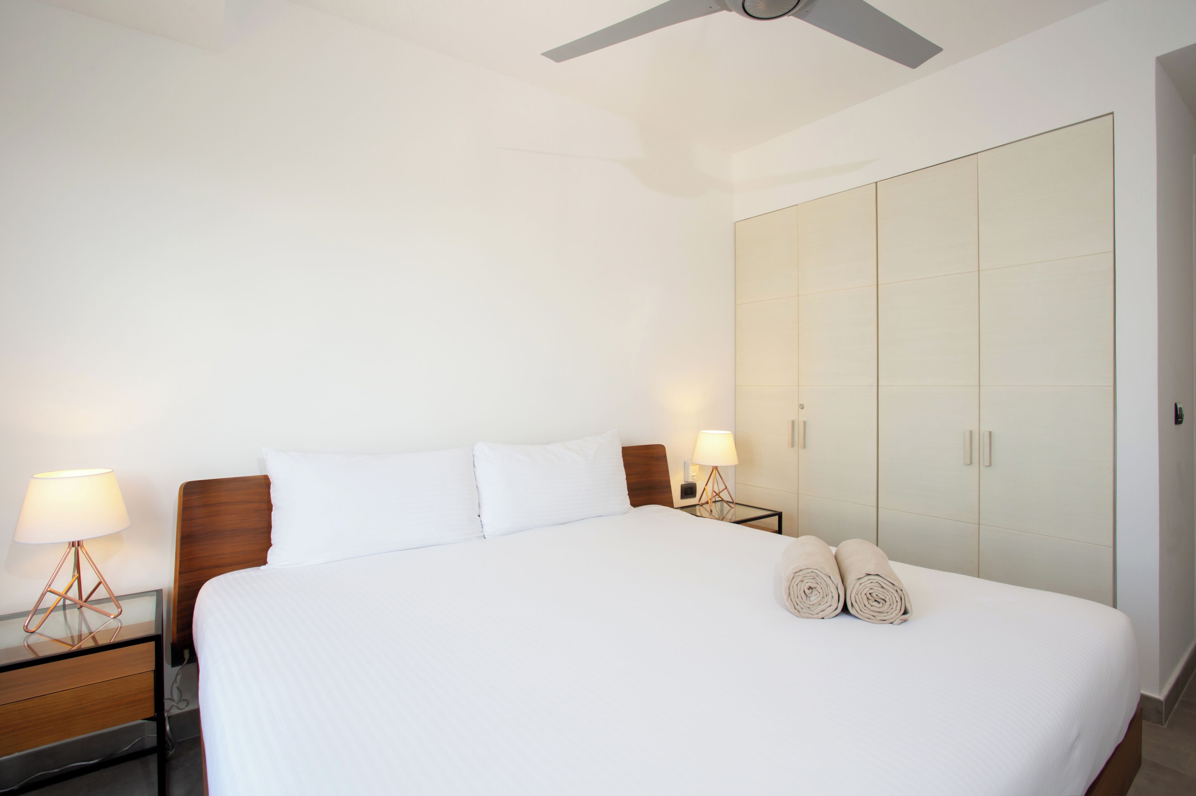 Quarto de hotel com uma cama grande, duas mesas de cabeceira com luminárias, um ventilador de teto e um guarda-roupa.