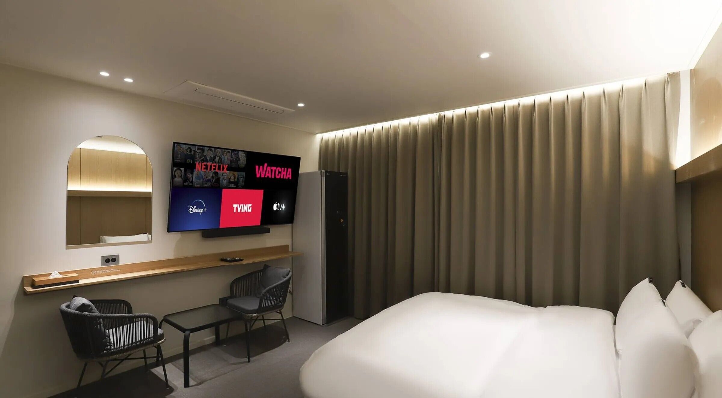 Quarto de hotel com cama, escrivaninha com cadeira, TV fixada na parede exibindo diversos serviços de streaming e uma janela grande com cortinas.