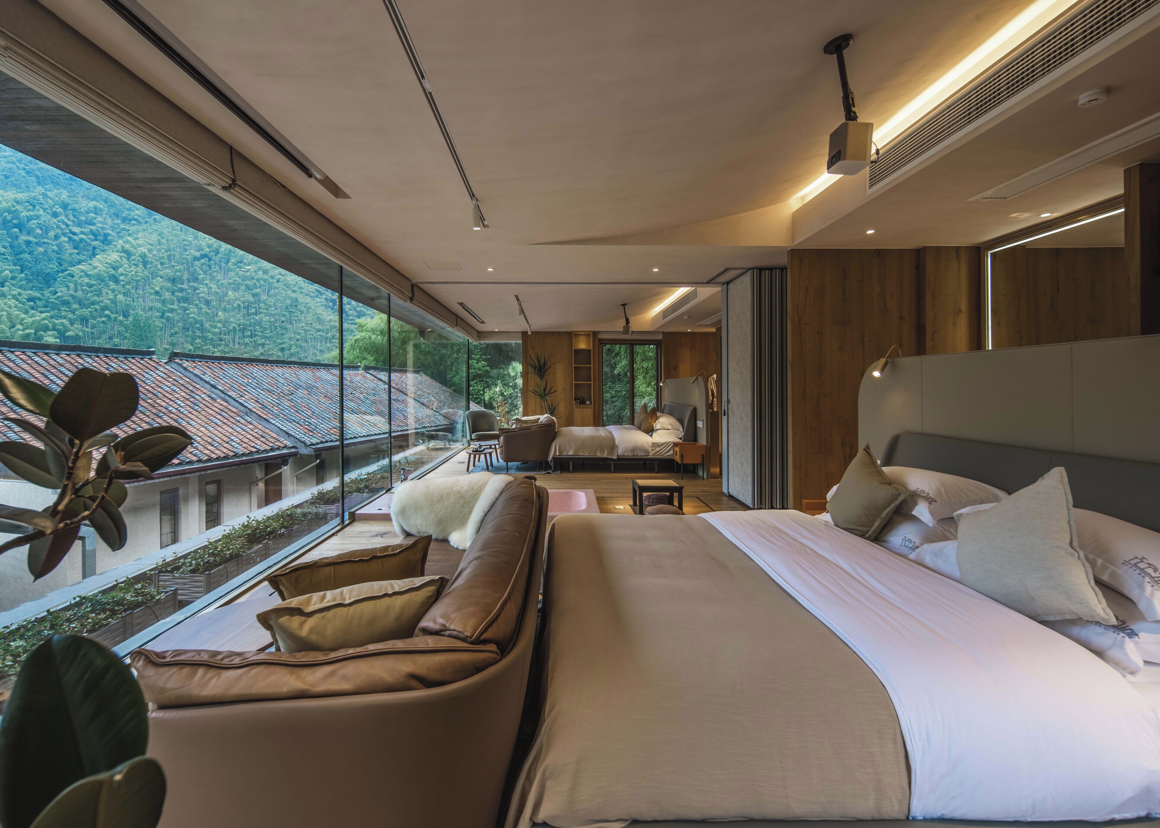 Quarto moderno com uma cama grande, um sofá e uma escrivaninha. Vê-se uma floresta através de grandes janelas.