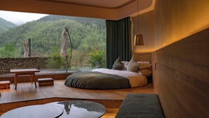Quarto moderno com piso de madeira, cama com travesseiros, um banco e uma mesa. Há vista de uma montanha arborizada por uma grande janela.